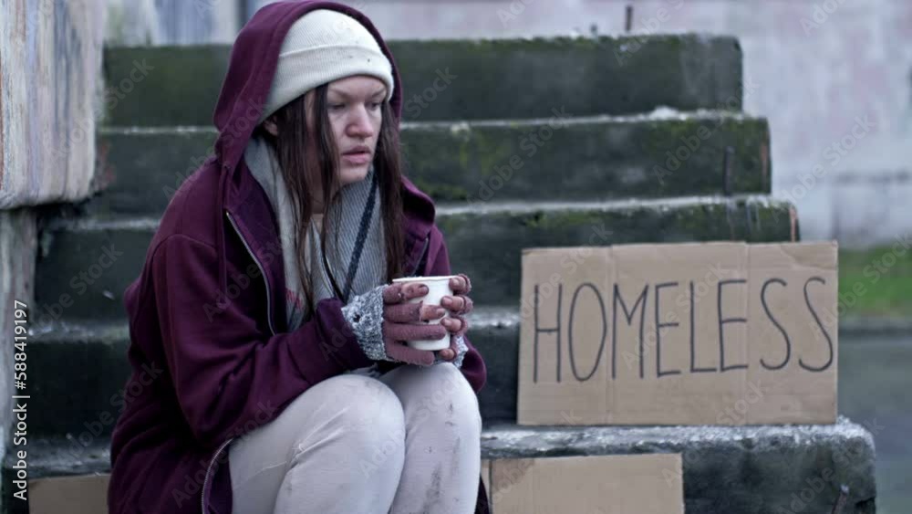 Homeless young woman is poorly dressed, dirty and needs help. Beside her is a handwritten poster ...