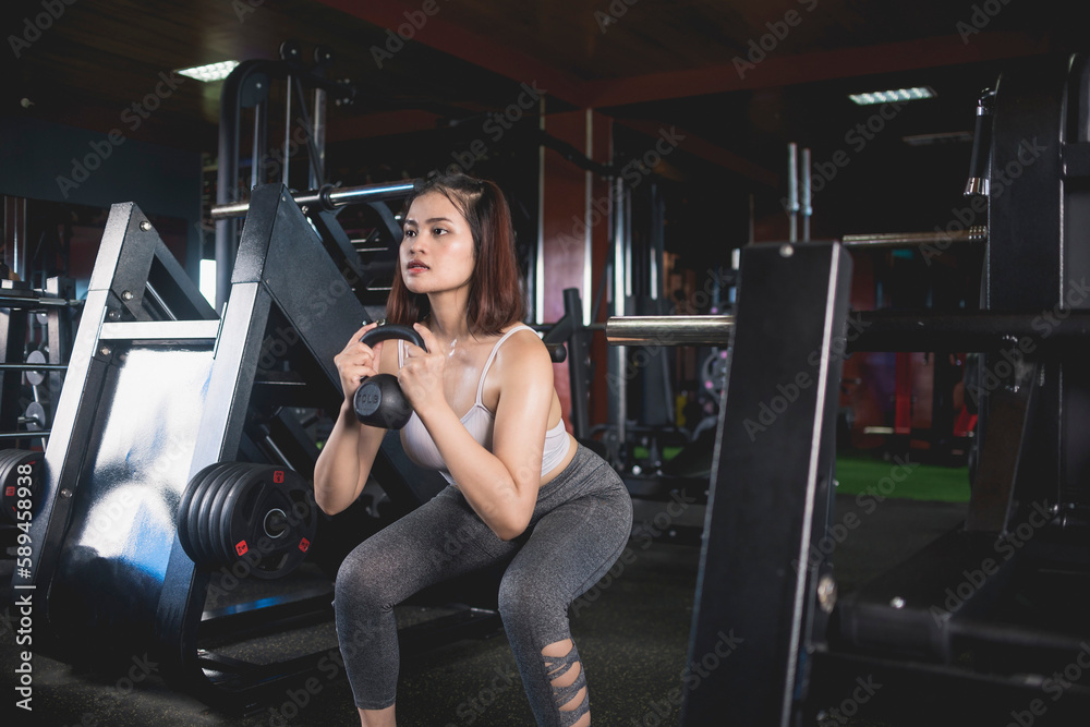 A young asian woman does a set of goblet squats with a kettlebell. Holding the weight in front ...