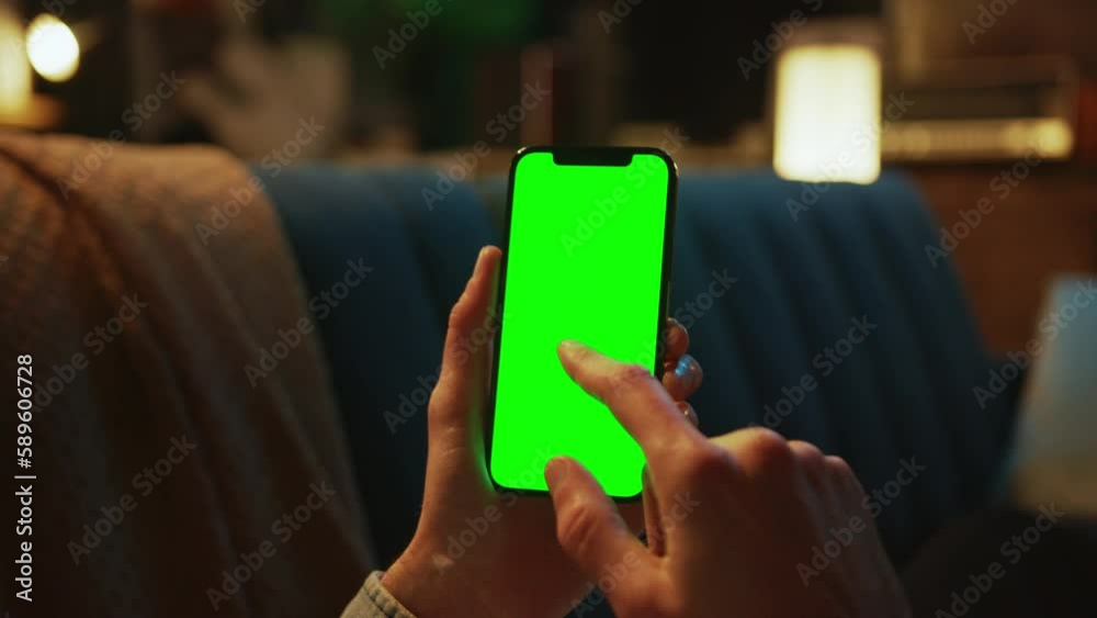 Close up hands holding and using mobile phone with a vertical green screen lying on a sofa ...