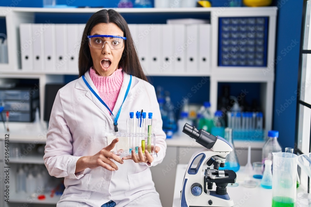 Young brunette woman working at scientist laboratory scared and amazed ...