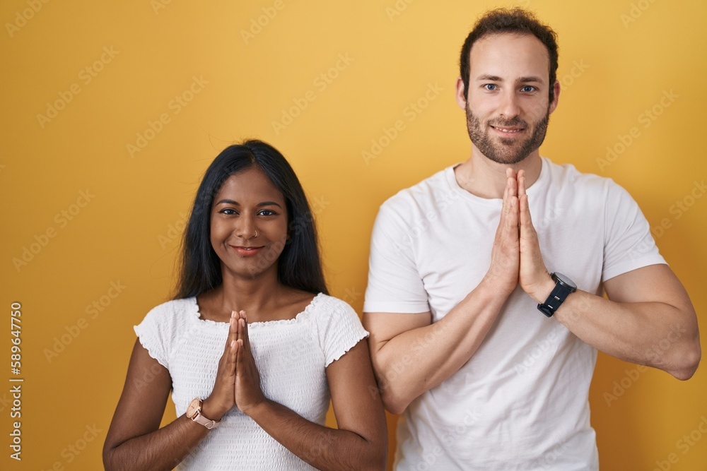 Interracial couple standing over yellow background praying with hands ...
