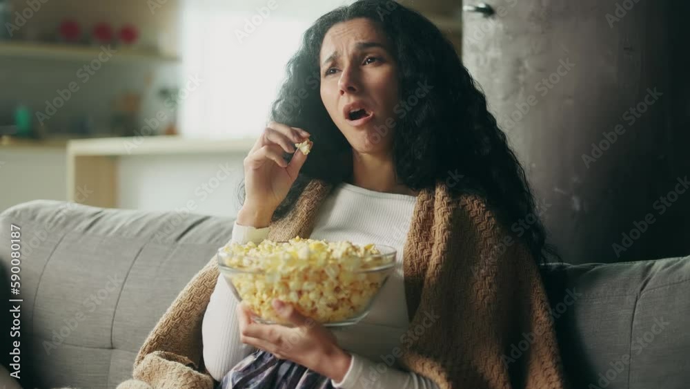 Portrait of sensitive young hispanic woman sits on sofa at home eating ...
