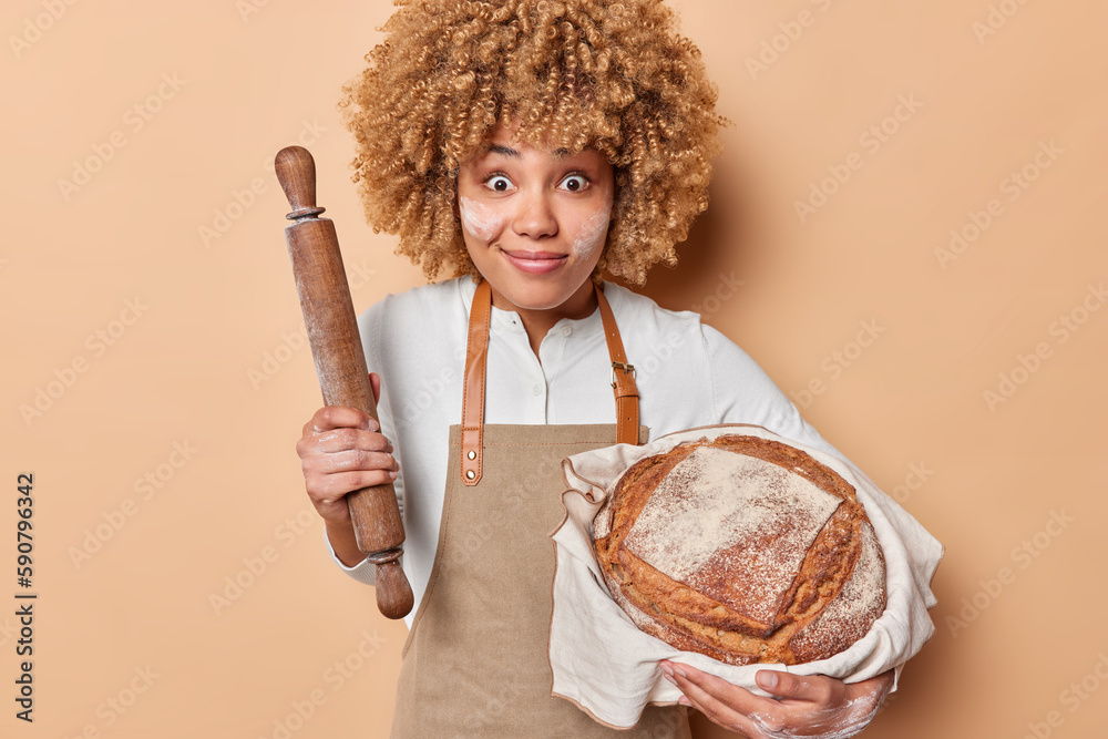 Stunned curly haired professional female baker holds wooden rolling pin ...
