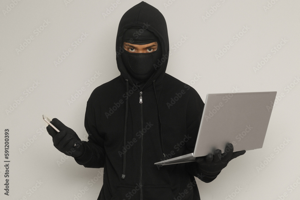 Portrait of mysterious man wearing black hoodie and mask doing hacking activity on laptop ...