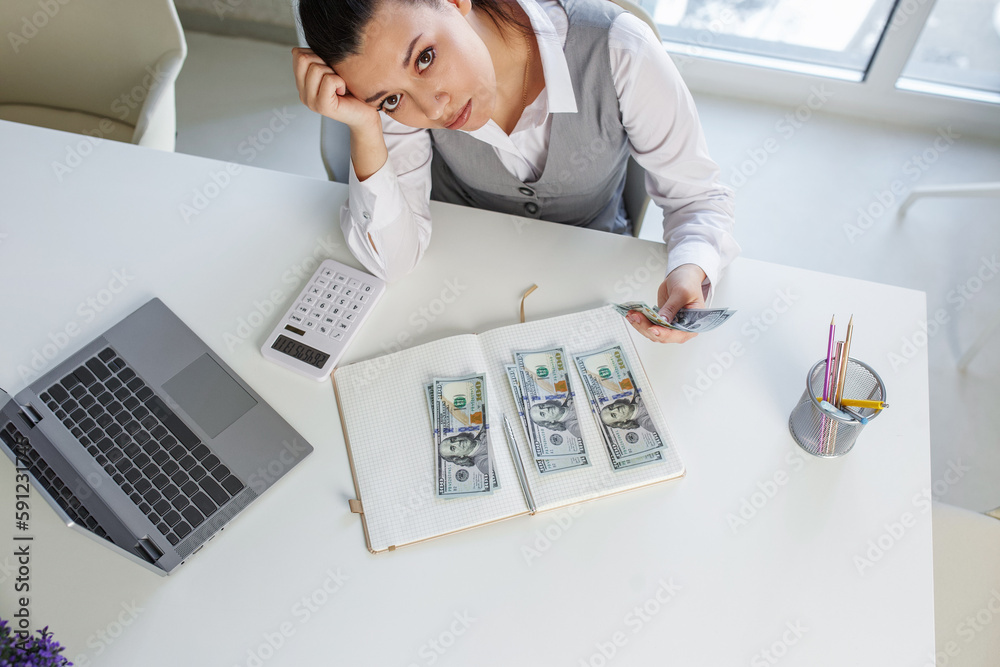 Sad business woman sits at work desk in front of laptop and counts cash ...