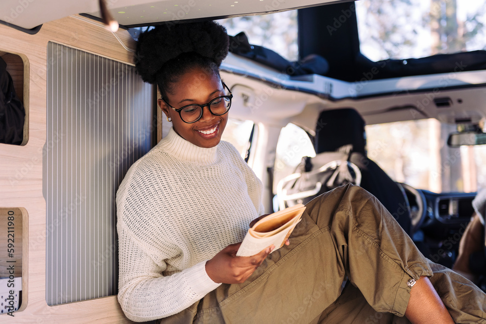 smiling black woman relaxing in camper van reading a book, concept of ...