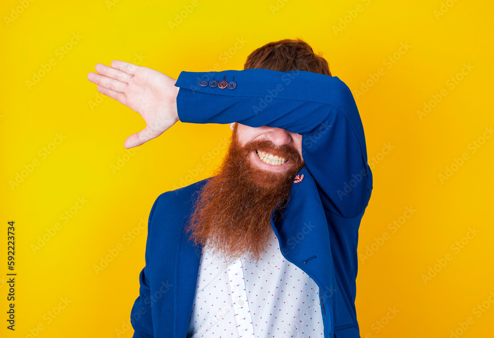 red haired man wearing blue suit over yellow studio background covering eyes with arm smiling ...