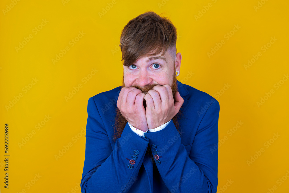 Fearful red haired man wearing blue suit over yellow studio background ...