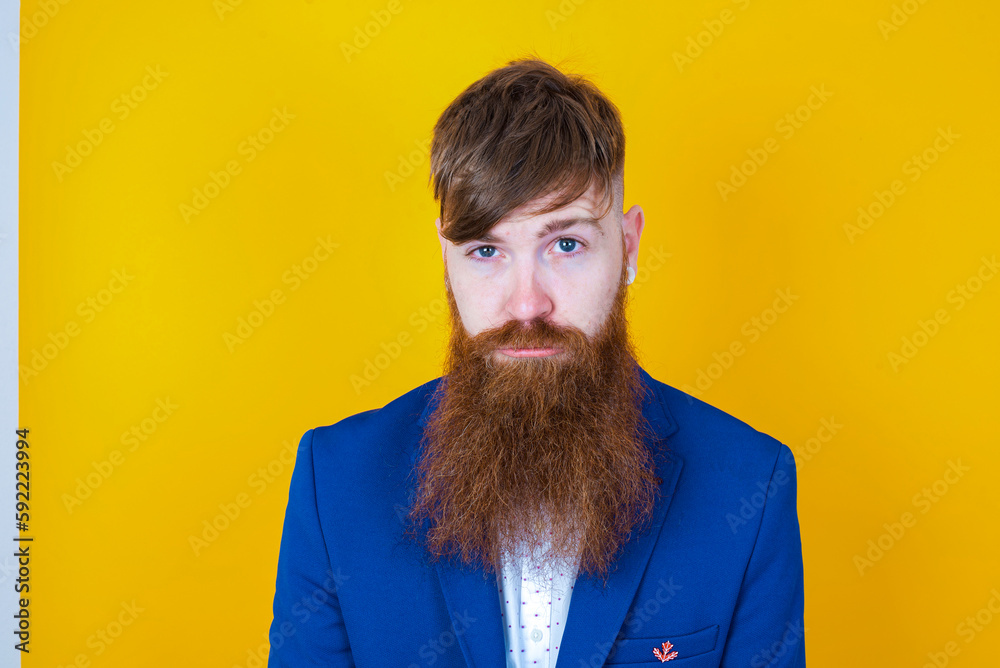 Displeased upset red haired man wearing blue suit over yellow studio background frowns face as ...