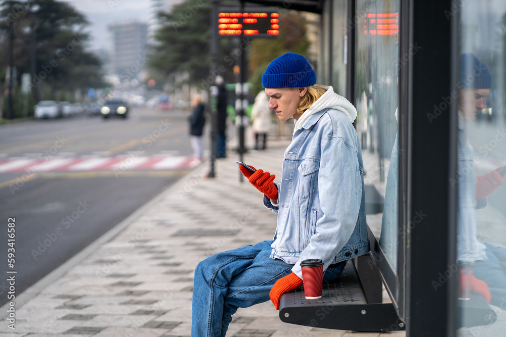 Stylish guy waiting bus sitting on bus stop bench in downtown with ...