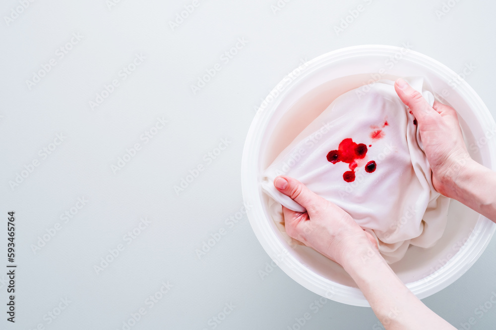 Female hands soak blood stain on the white clothes in a plastic basin ...