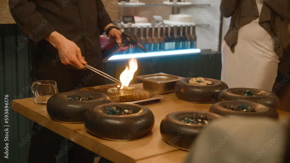 Male chef prepares meal in flambe style in front of people in modern ...