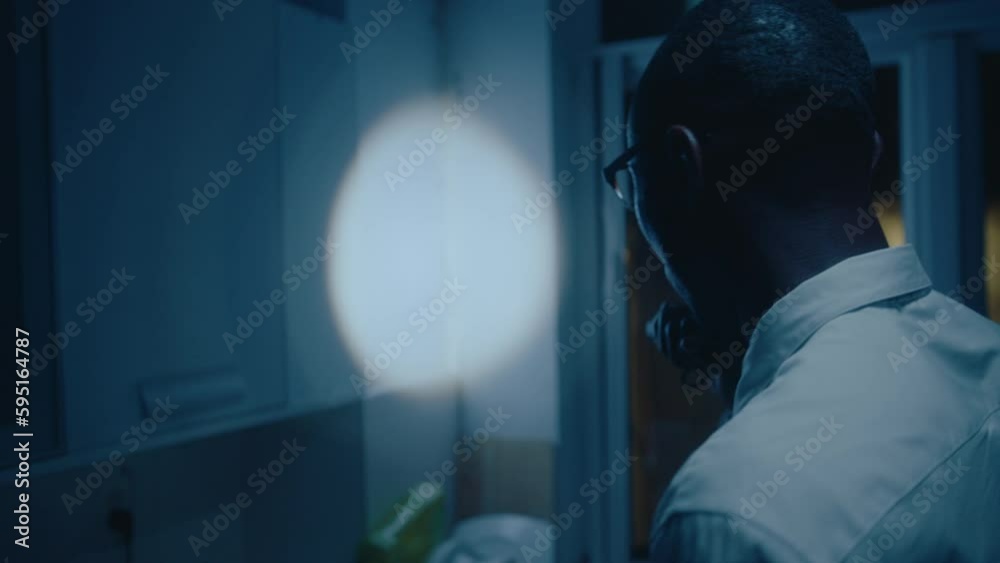 Black investigator inspecting wall cabinets with flashlight in dark ...