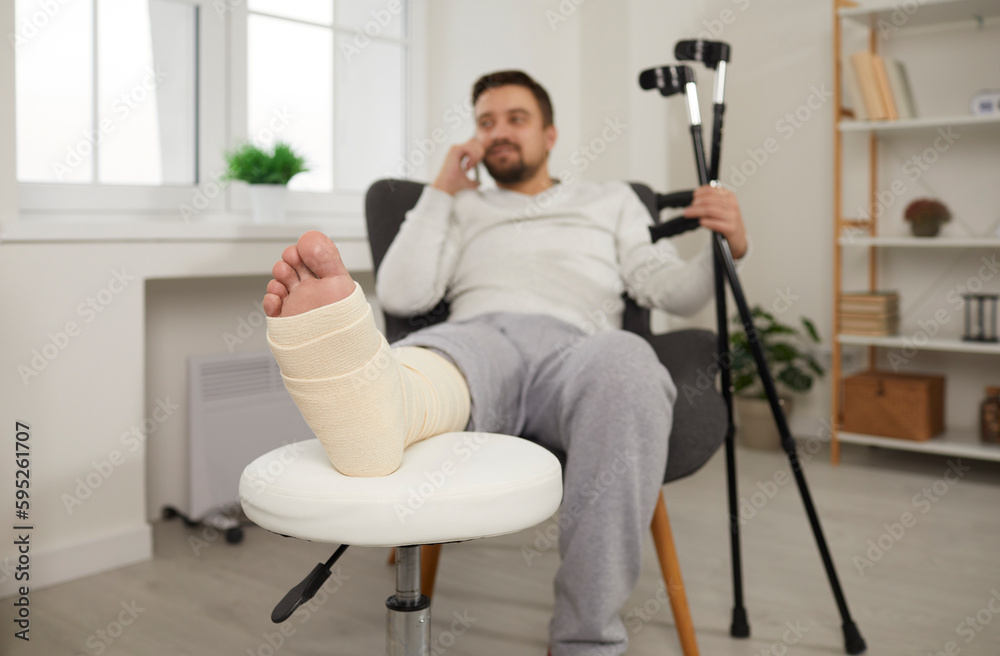 Unhealthy young man is sitting at home in chair with broken leg in cast ...