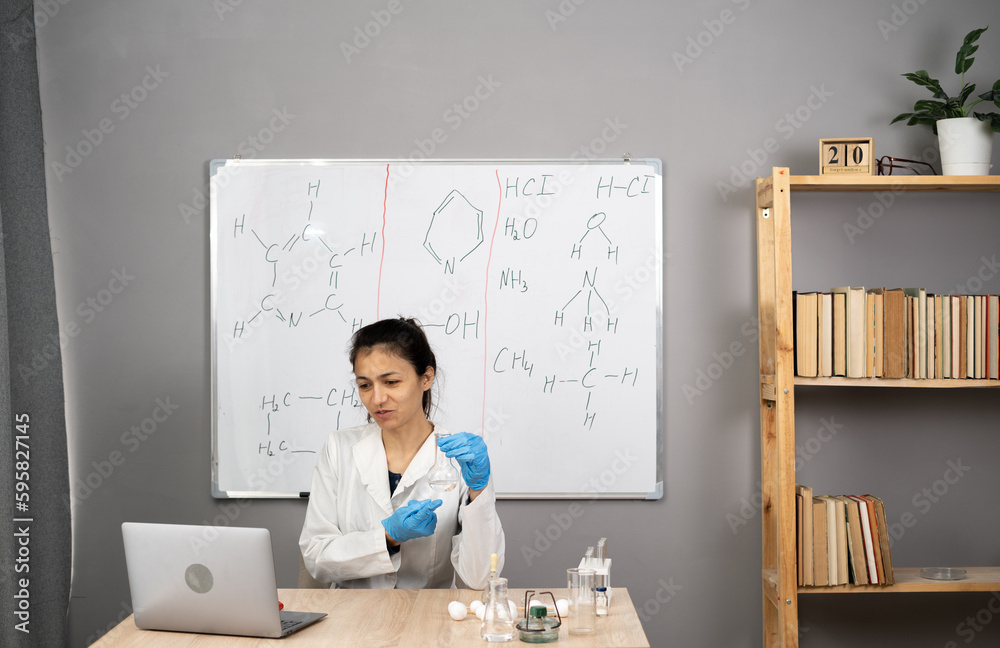 Smiling female chemistry teacher wearing white coat with laptop ...