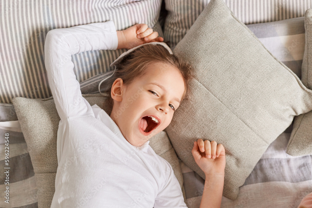 Sleepy little girl yawning in the morning while lying in bed on pillows, keeps mouth open lazy ...
