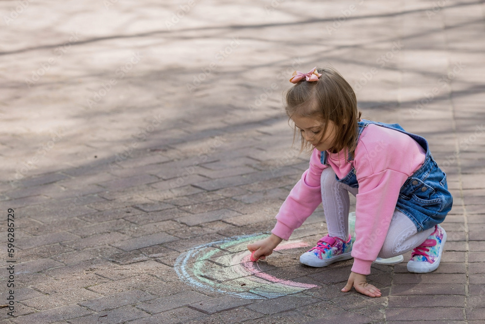 A happy girl in pink clothes draws a rainbow with chalk on the asphalt ...