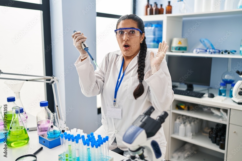 Young hispanic woman working at scientist laboratory scared and amazed ...