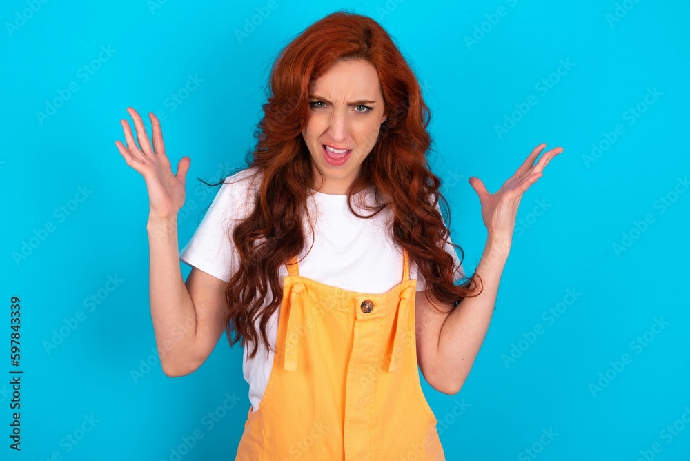 Crazy outraged Young redhead woman wearing orange overall over blue ...