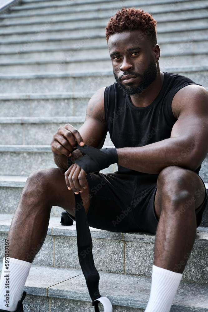 Fit sporty young black man sitting at urban stairs wrapping hands with ...