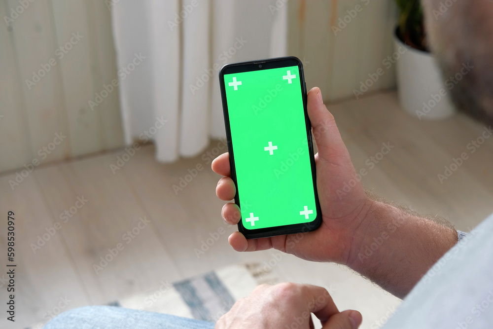 Young man sitting at home holding smartphone green mock-up screen in hand. Male person using ...