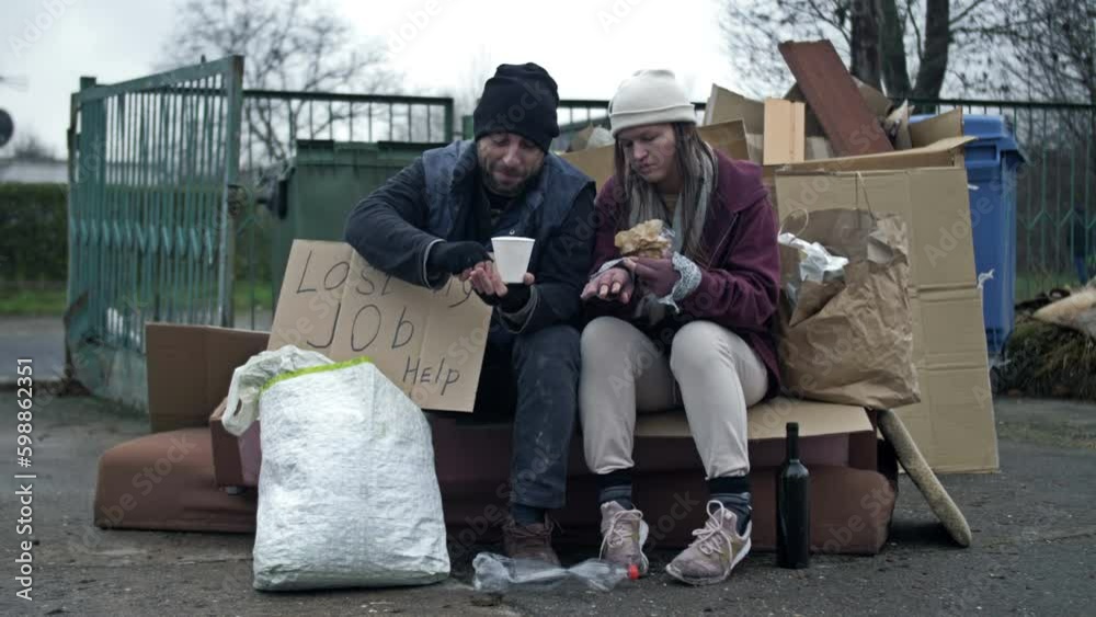 Two dirty and poorly dressed homeless people, a man and a woman, sit by ...