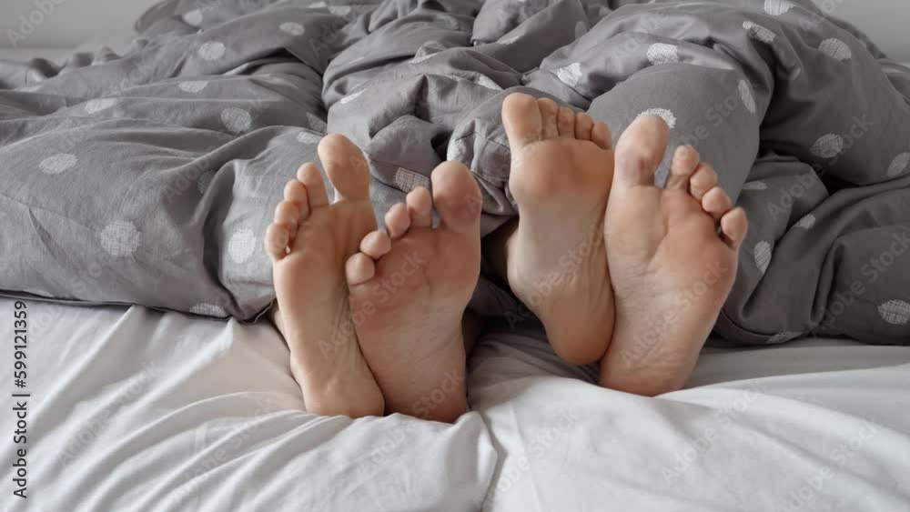 Closeup as the feet of a man and a woman lie entwined under the blanket ...