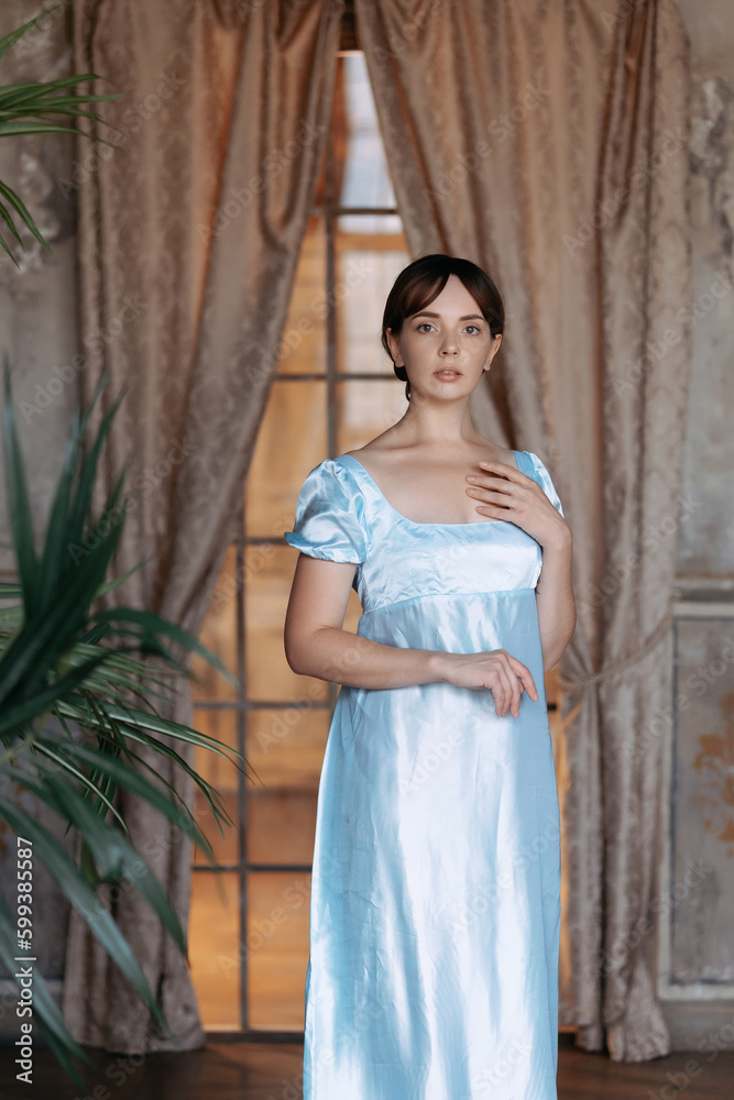 A young beautiful girl in a blue Regency dress stands at a wooden ...