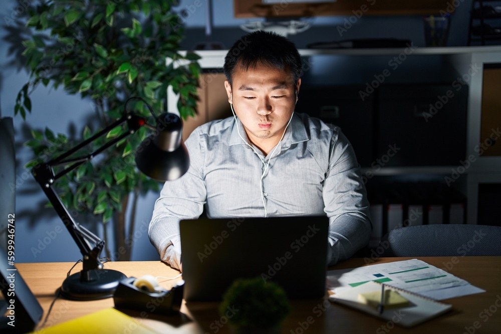 Young chinese man working using computer laptop at night puffing cheeks ...