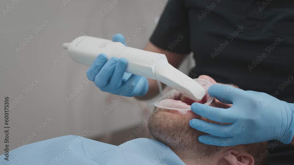 A male orthodontist doctor scans a man's teeth and jaw in the clinic's ...