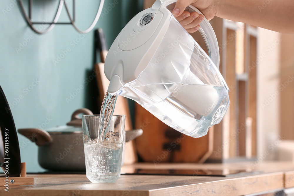 Woman pouring water from modern filter jug into glass on kitchen ...
