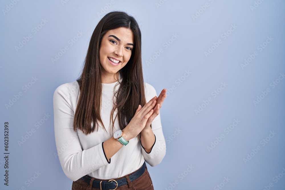 Young brunette woman standing over blue background clapping and ...