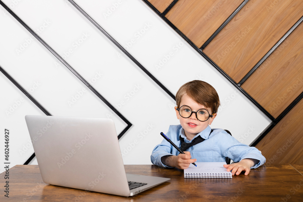 Little boy in round eyeglasses, blue shirt and tie sitting at the desk ...