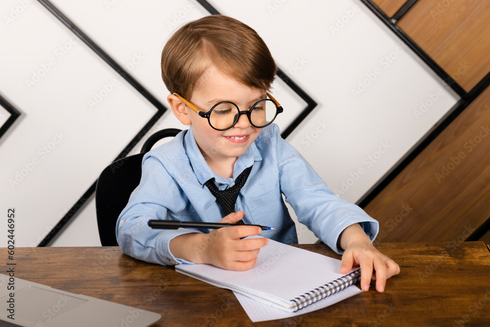 Little boy in round eyeglasses, blue shirt and tie sitting at the desk ...