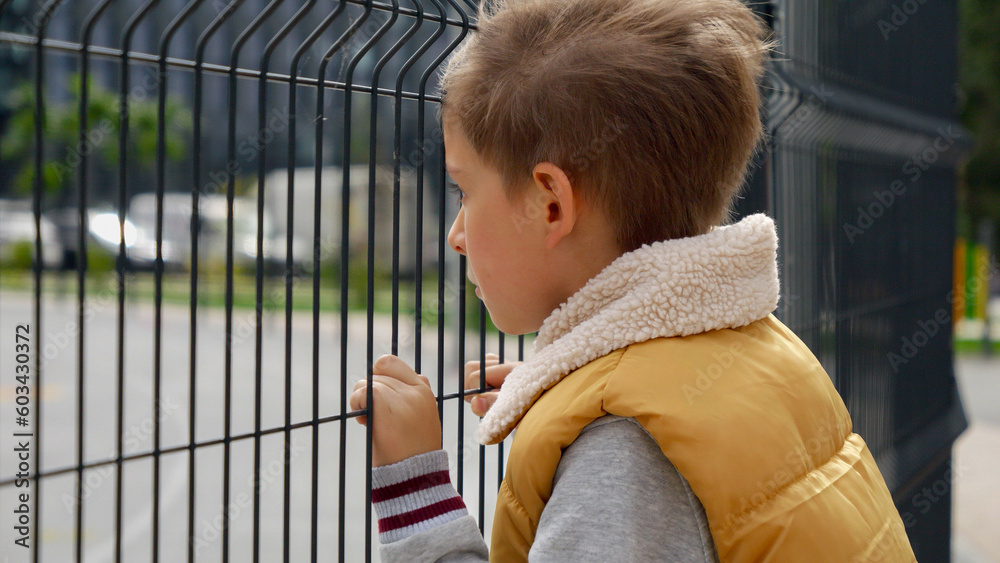 Portrait of upset little boy feeling alone looking through metal fence ...
