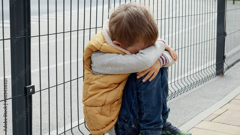 Lonely boy feeling alone on playground sitting next to metal fence and ...