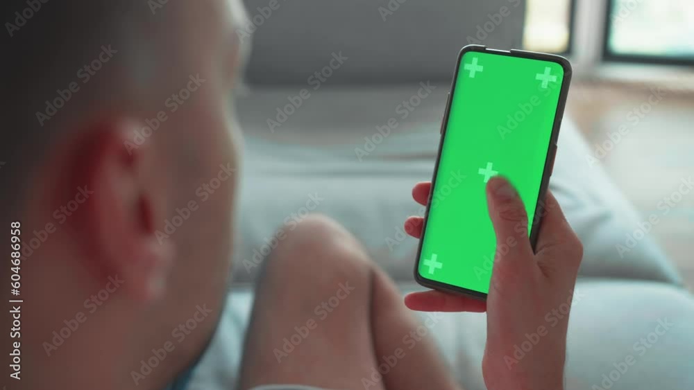 Young Caucasian Man at Home Lying on a Couch using Smartphone swiping with Green Mock-up Screen ...