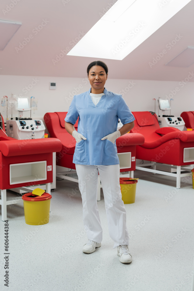 Positive Multiracial Nurse In Latex Gloves Standing With Hands In Pockets Of Uniform Near