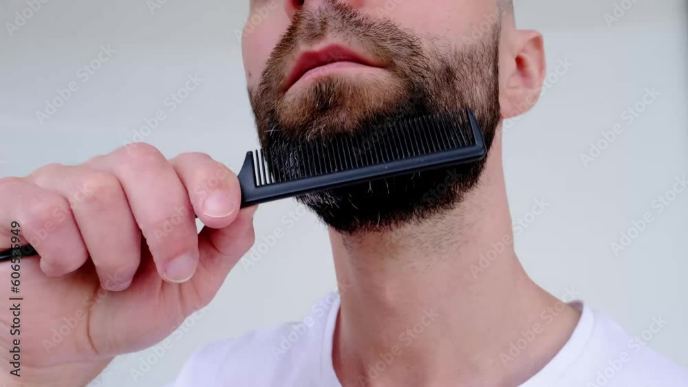 man combing his well-groomed beard with fine plastic comb, close-up part of the face, bearded ...