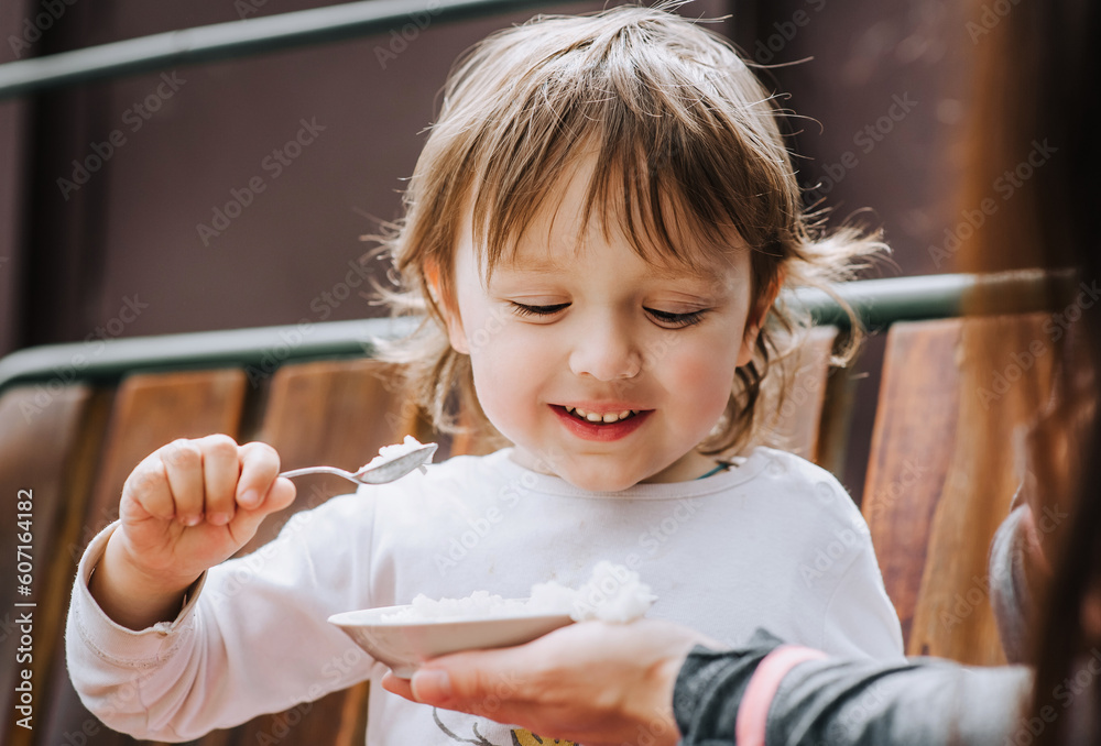 A beautiful little boy, a happy child, eats rice porridge with a spoon ...