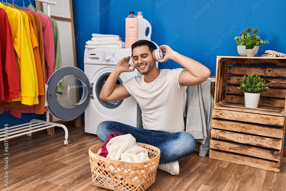 Young hispanic man listening to music washing clothes at laundry room ...