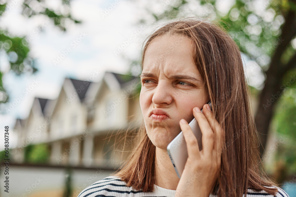 Unhappy sad brown haired young woman talking with cellphone frowning ...