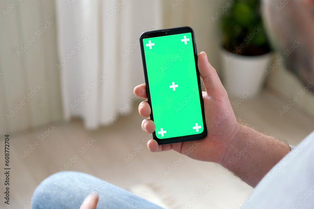 Hipster man sitting at home holding smartphone green mock-up screen in ...
