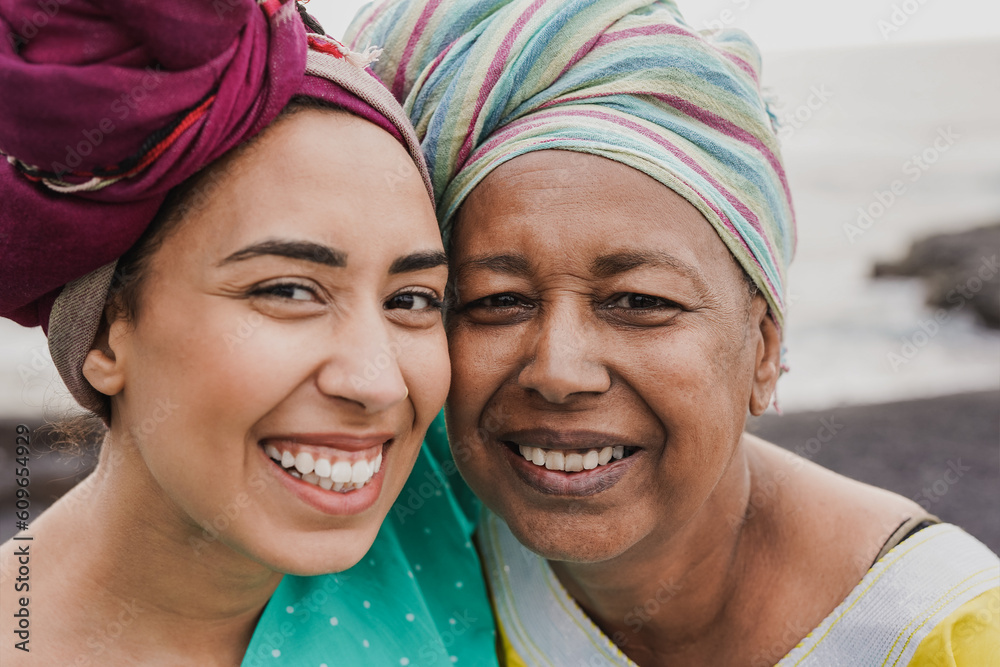 Happy african mother and adult daughter smiling on camera with beach in the background while ...