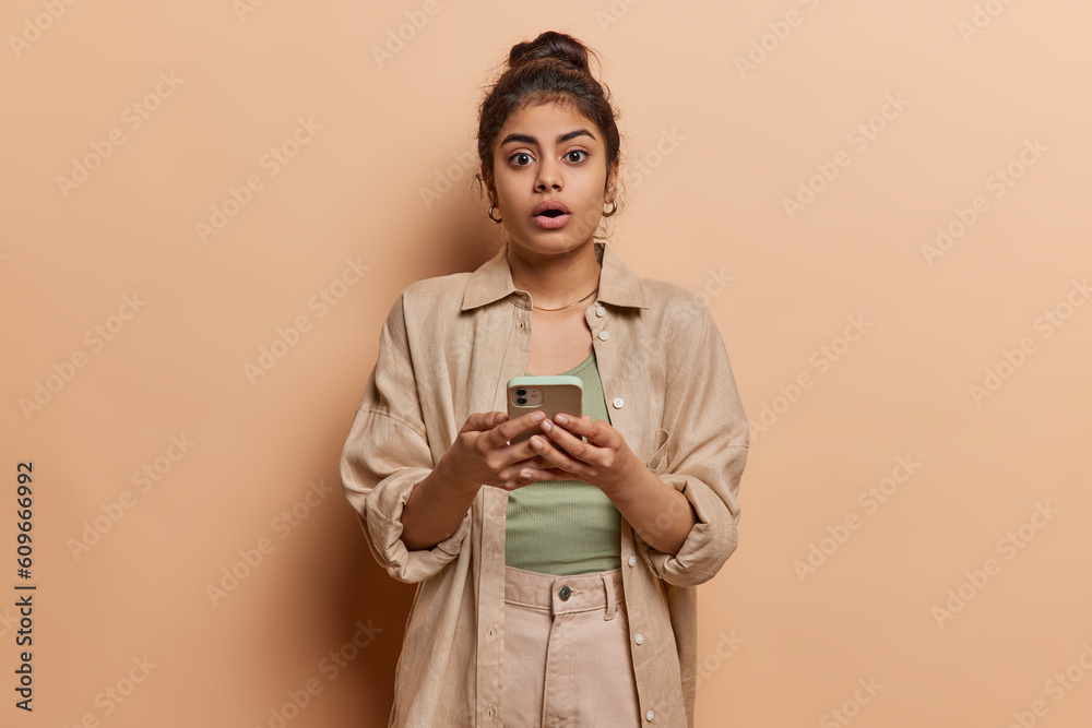 Horizontal shot of impressed young Indian woman poses with cellphone ...
