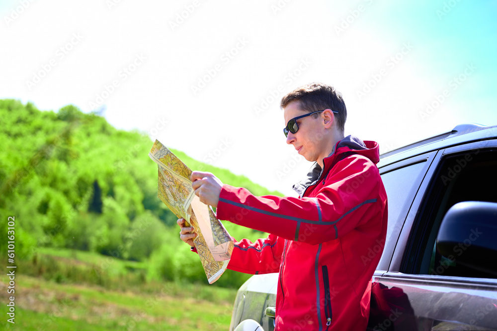 A man leans against a car, holding a map in his hands, as he looks for directions. The traveler ...