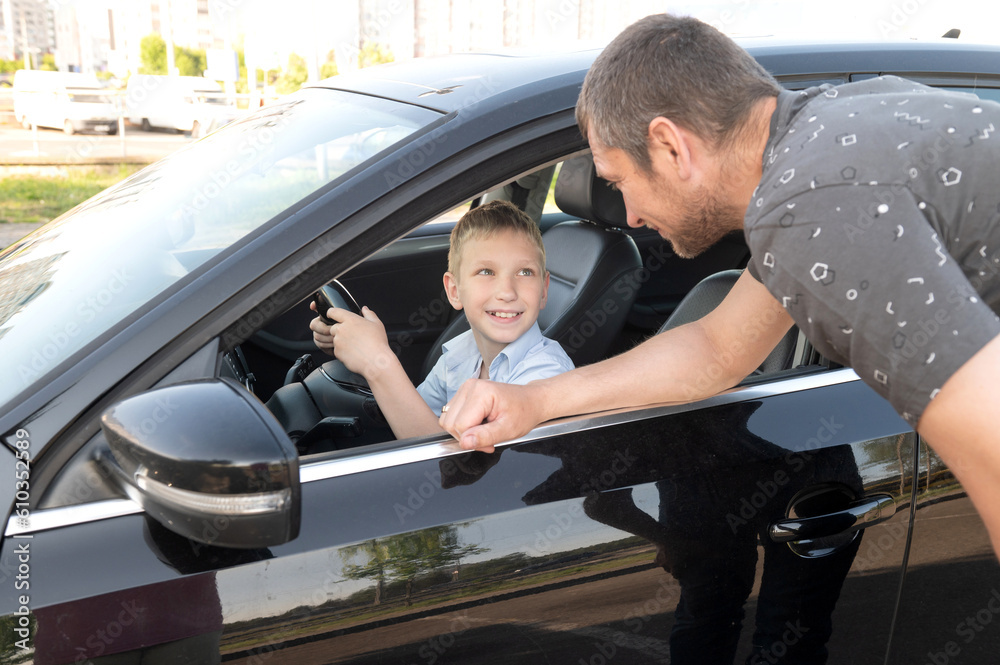 Cute boy is sitting at the wheel in the car, dad is looking out the car ...