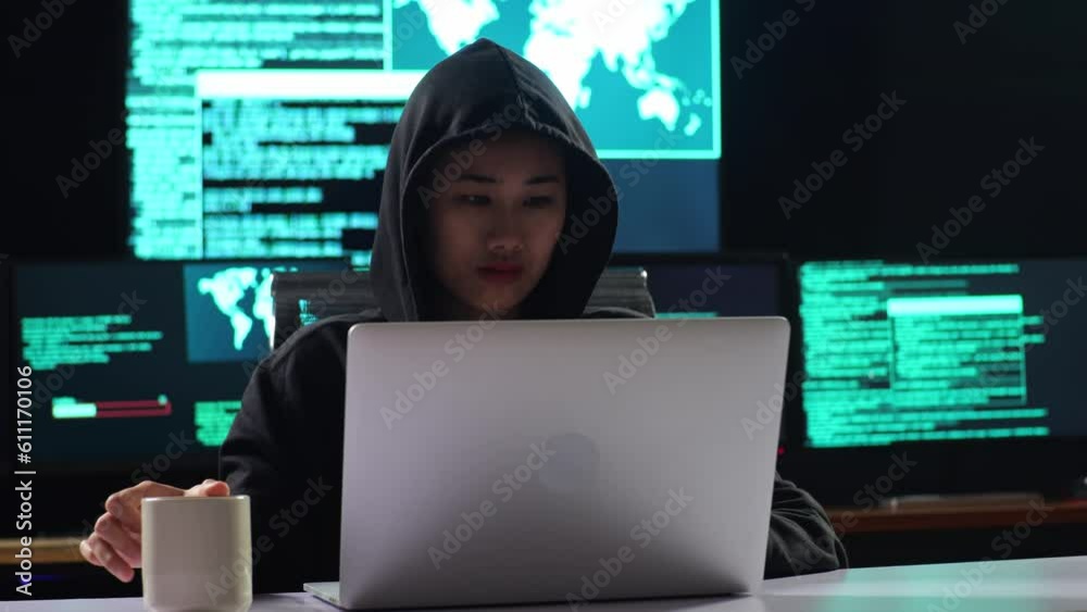 Asian Female Hacker In A Hood Drinking Coffee While Works On A Laptop With Maps And Data On ...