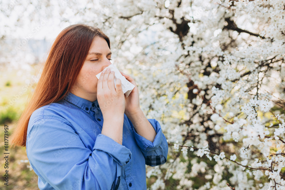 Sneezing young redhead woman with nose wiper among blooming trees in ...