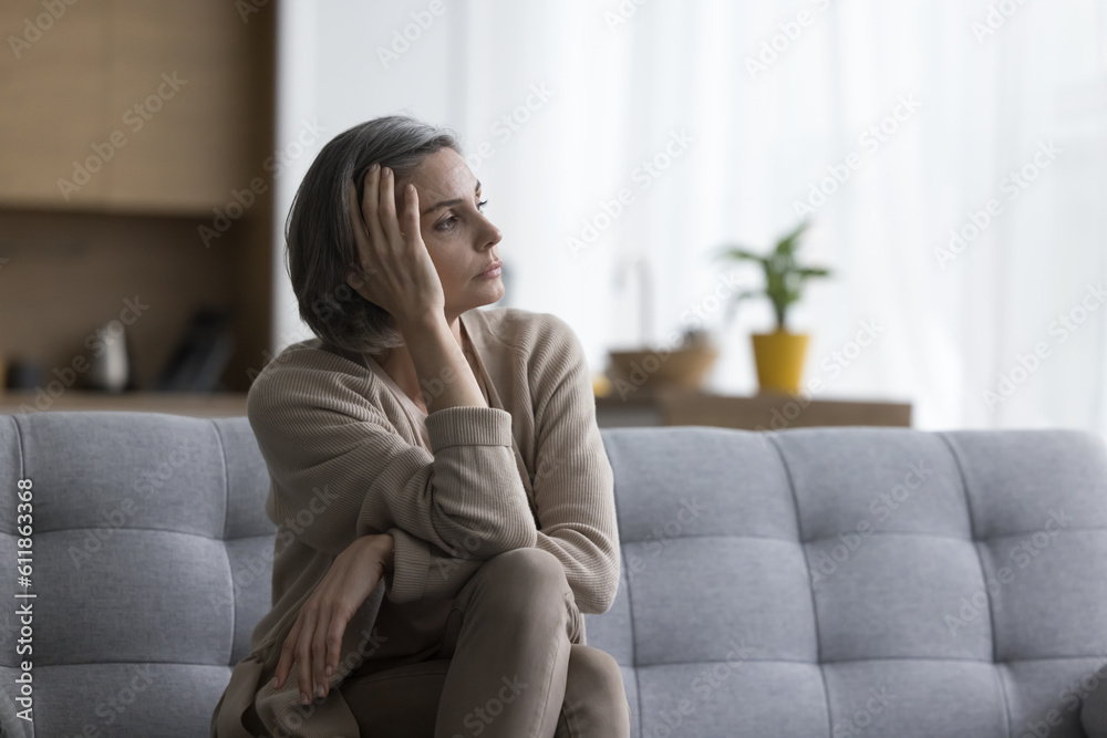 Sad thoughtful grey haired woman sitting on sofa at home, looking at ...